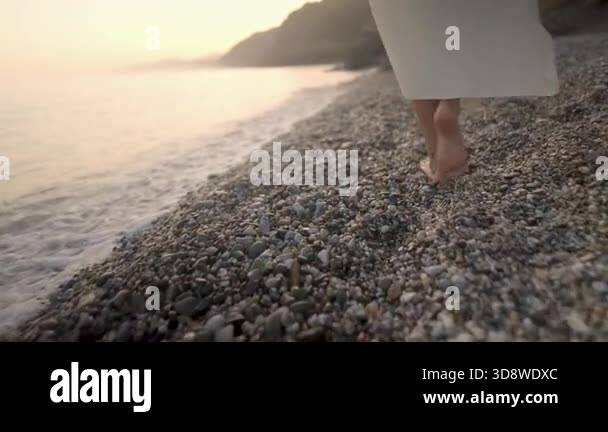 Barefoot woman in white dress walking along pebble beach during golden ...