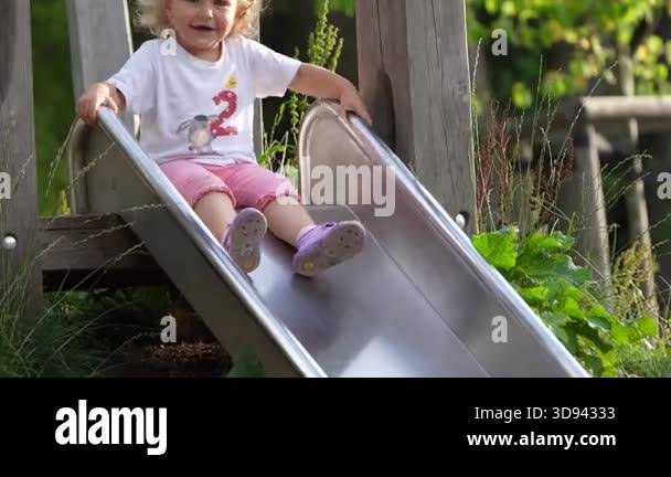 A sweet little girl enjoys playing on a metal slide in a park. She is ...