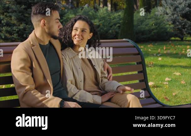 Cheerful hispanic couple in autumn park talking happy relaxing on bench ...