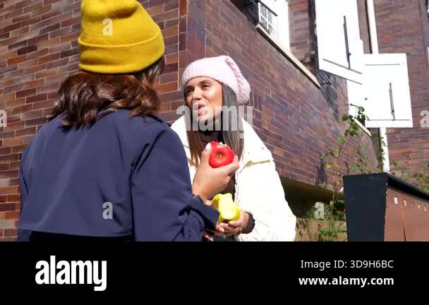Two cheerful female friends standing by a brick wall, eating apples and ...