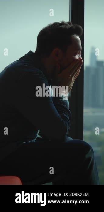 man seated by office window holding face in hands, city skyline ...