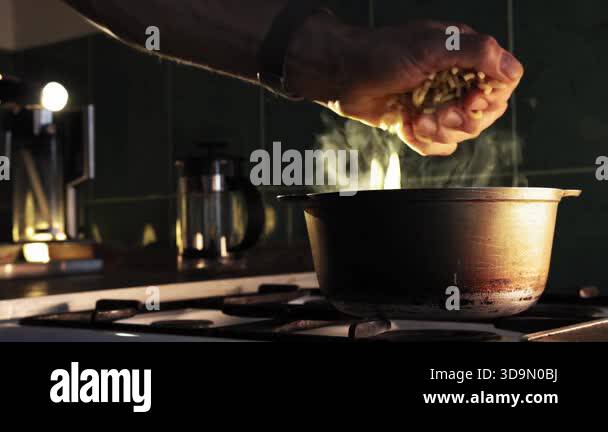 In a dark kitchen, a man pours pasta from his hand into a pot of ...