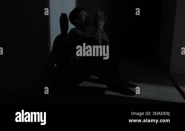 Frightened man sitting on the floor in the dark with trembling hands ...