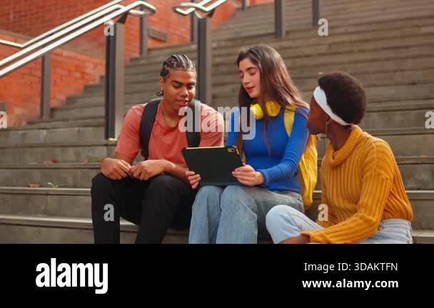 Three multicultural friends sitting on campus stairs discussing a ...