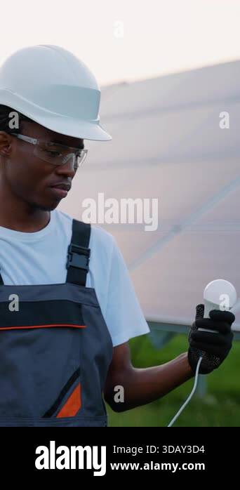 Young black technician in a hard hat holding a glowing led light bulb ...