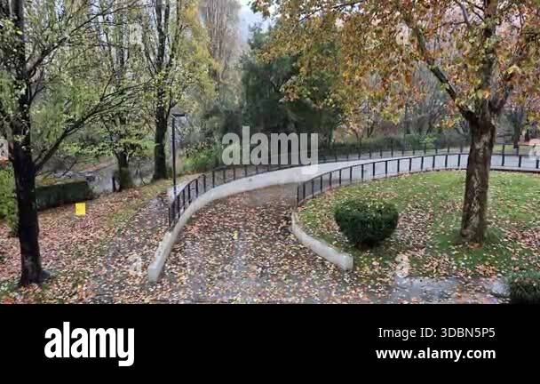 A quiet park pathway curves through autumn trees on a rainy day, with ...