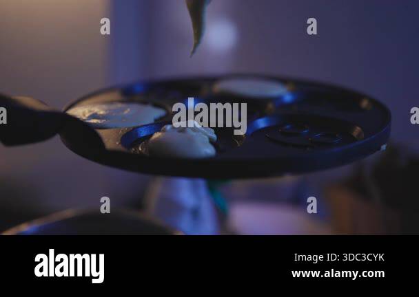 Thick batter flows from above into circular molds of a stovetop pan ...