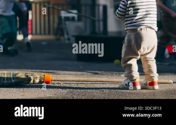 Toddler standing near fallen broom watching older children run and play ...
