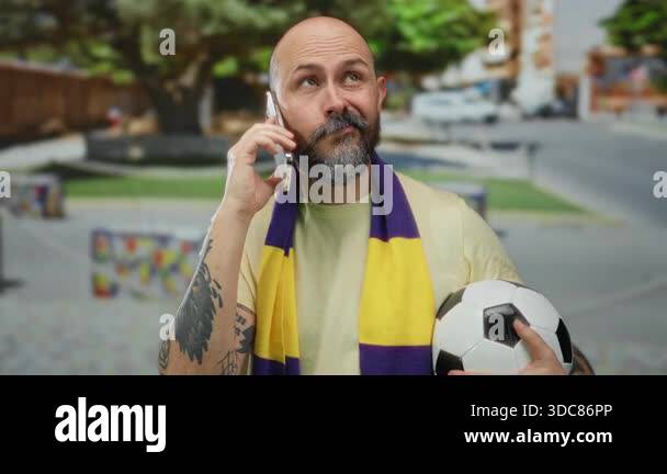 Caucasian bald man with beard holding soccer ball talks on smartphone ...