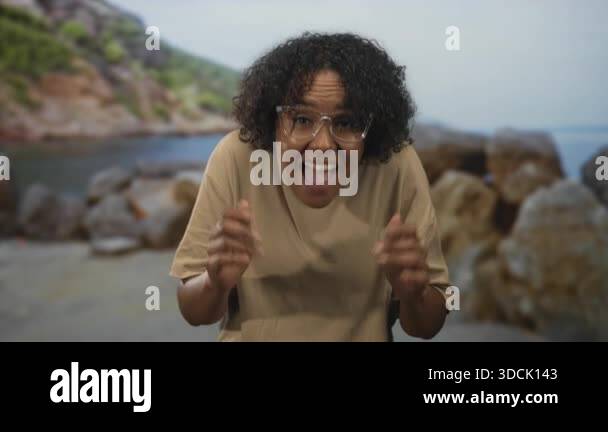 Young woman touches cheeks and adjusts glasses on rocky beach at sunset ...