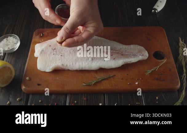 A hand sprinkles spices over a raw fish fillet placed on a wooden ...