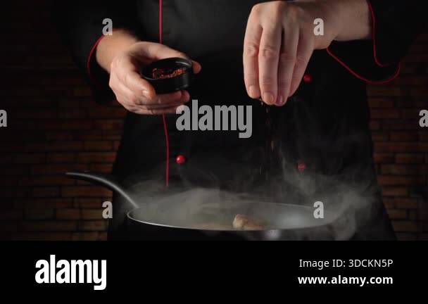 A person in a dark chef outfit sprinkles spices into a pan on a ...