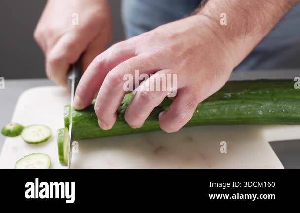 A hand slices a fresh cucumber on a white cuttingboard with a sharp ...