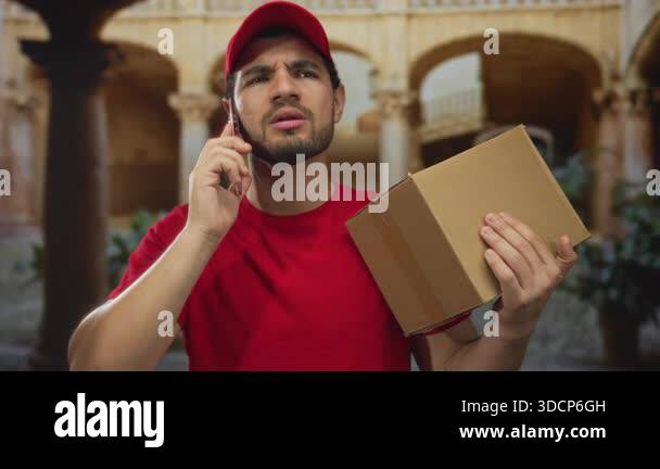 Young delivery man in red shirt and cap holds cardboard box while ...
