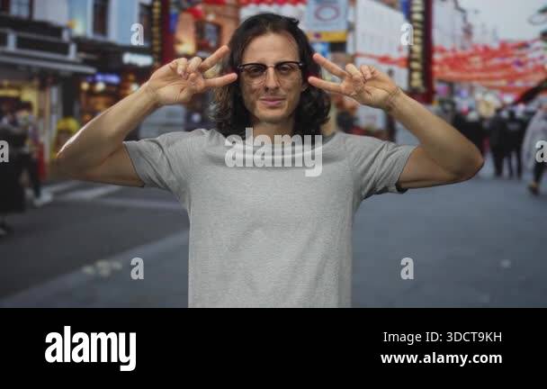 Man holding up two peace signs with both hands on a street wearing grey ...