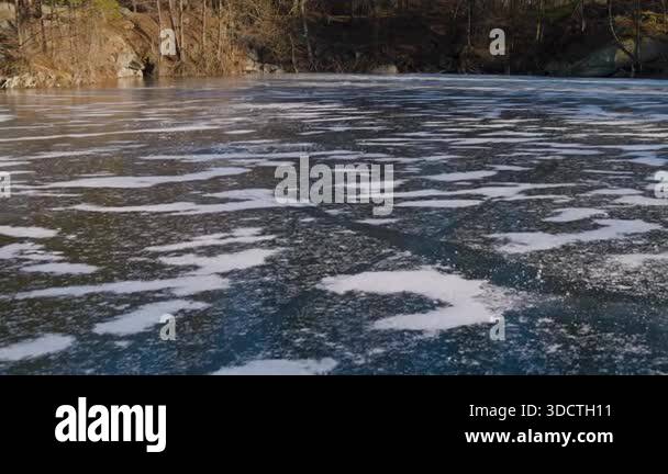 Frozen lake with cracked ice and snow patches leafless trees and rocky ...