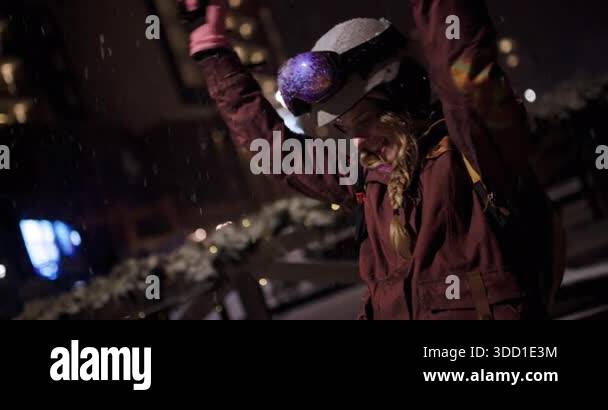 Happy snowboarder girl dancing at night in a ski resort under snowfall ...