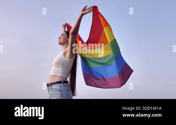 A smiling young woman holds the Rainbow Pride Flag above her head in a ...