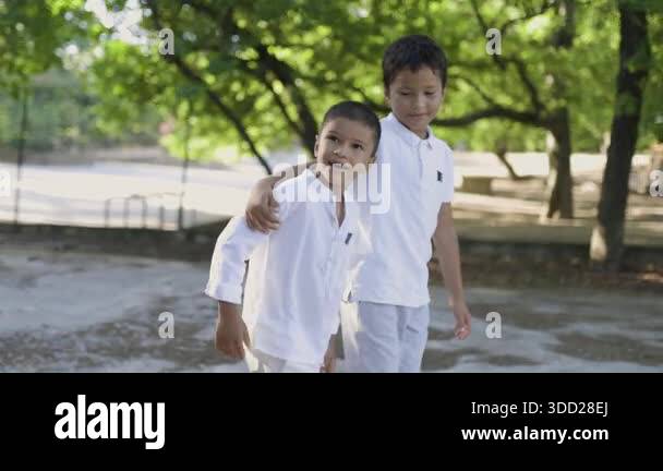 Two young brothers, wearing white shirts, walk together through a park ...
