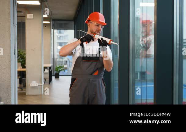 Professional builder in uniform and hard hat applying silicone sealant ...