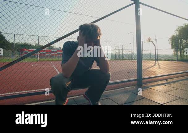 A teenage boy crouches near a fenced sports court, holding his head in ...