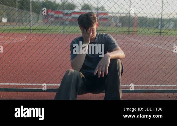 A frustrated teenage boy crouches beside a chain-link fence near a ...