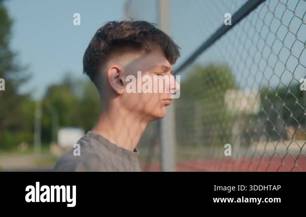 A teenage boy stands beside a chain-link fence, staring forward in ...