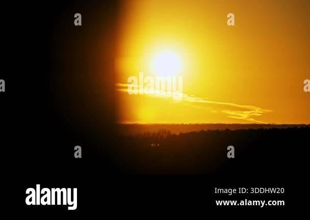 Bright golden sun sets over a silhouetted treeline with a wispy cloud ...