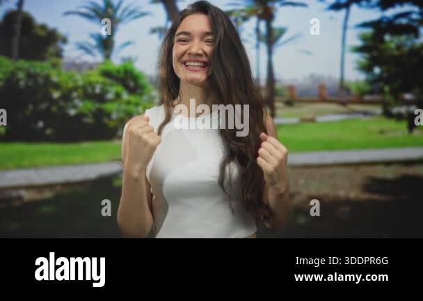 Woman clenches fists and smiles under sunlight amid palm trees in green ...