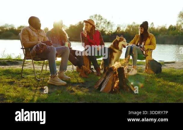 Sunny attractive friends sitting near campfire in nature. Young people ...