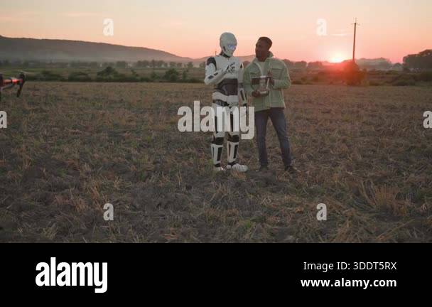 A human and robot work together in a scenic field at sunset ...