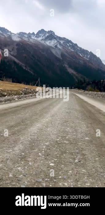 Man walking gravel mountain road under overcast sky toward snowcapped ...