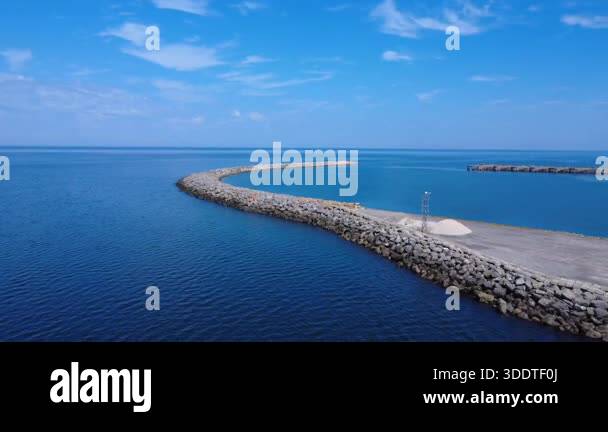 A long stone breakwater juts out into the calm blue sea, forming a ...