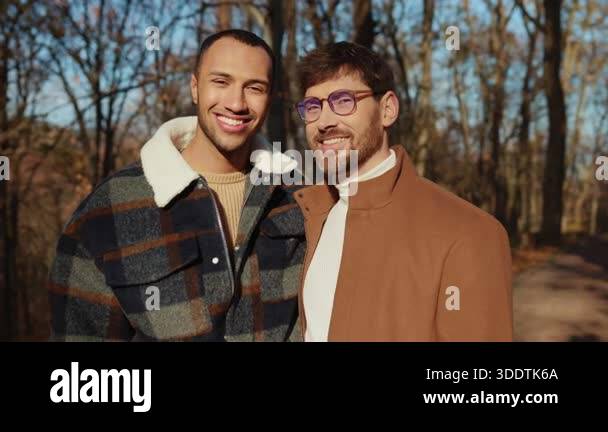 Two joyful young hugging European men in overcoats looking at camera ...