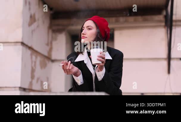 Young woman in red beret stands on street sipping coffee and slowly ...