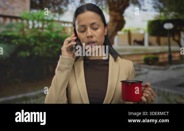 Woman talking on phone holding red cup in green park setting, wearing a ...