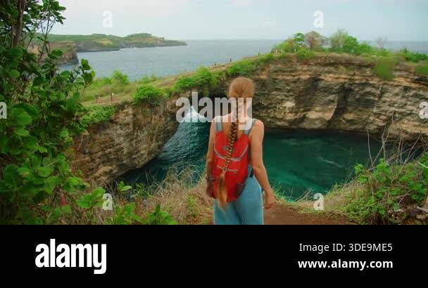 Young woman with long braid walking on path toward coastal arch at ...