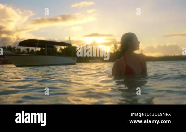 At sunset, a woman in a red bikini swims in the calm ocean waves ...