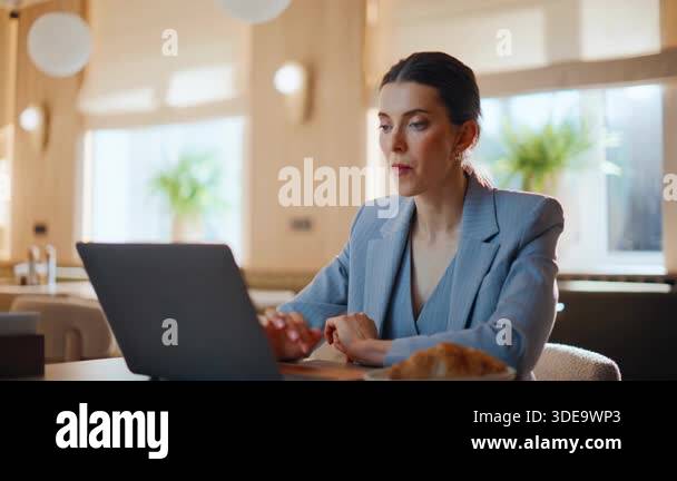 Confident businesswoman working laptop eating croissant at city cafe ...