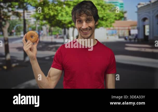 Man in red shirt holds glazed donut with left hand in sunny city street ...