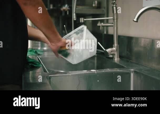 Male kitchen worker washing a large plastic food container in a ...