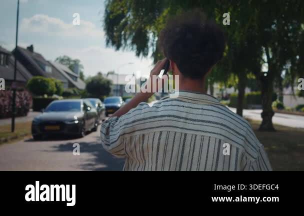 Young man on phone walking past parked cars on suburban street, striped ...