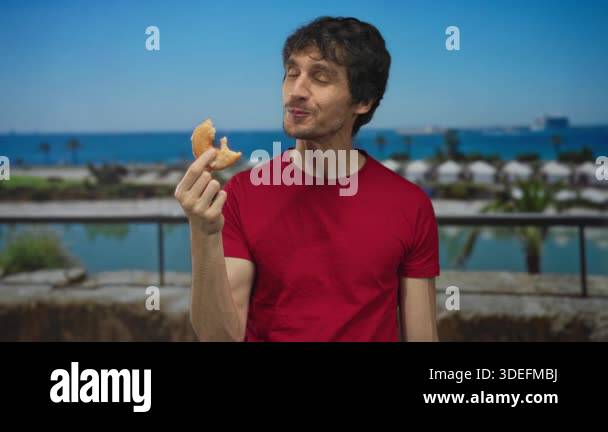 Man holds bitten sugar coated donut by hand at sunlit port overlooking ...