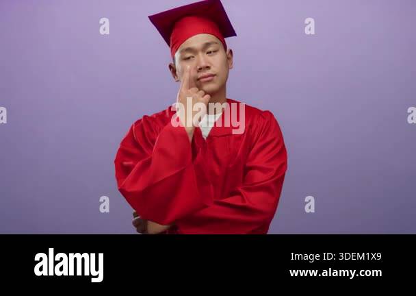 Young graduate man in red cap and gown touches his eye against a purple ...