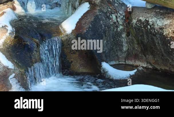 A miniature waterfall cascades over snow-covered rocks, creating a ...