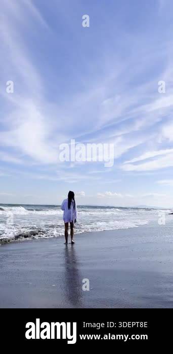Woman in white shirt walking slowly along the shoreline with smartphone ...