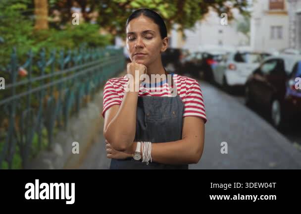 Woman with hand on chin on city street wearing denim apron and red ...