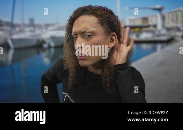 Transgender woman cups hand to ear beside a docked boat at a sunny port ...