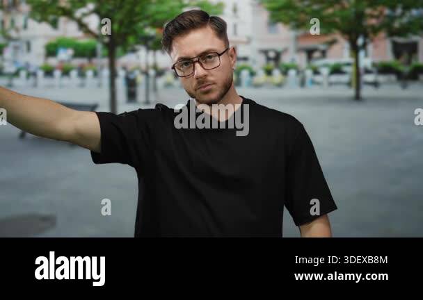 Young man with beard wearing glasses gestures on city street backdrop ...