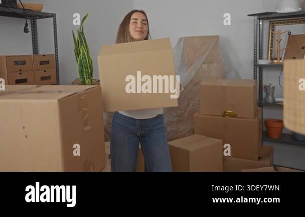 Young woman moving boxes indoors surrounded by packing supplies in new ...